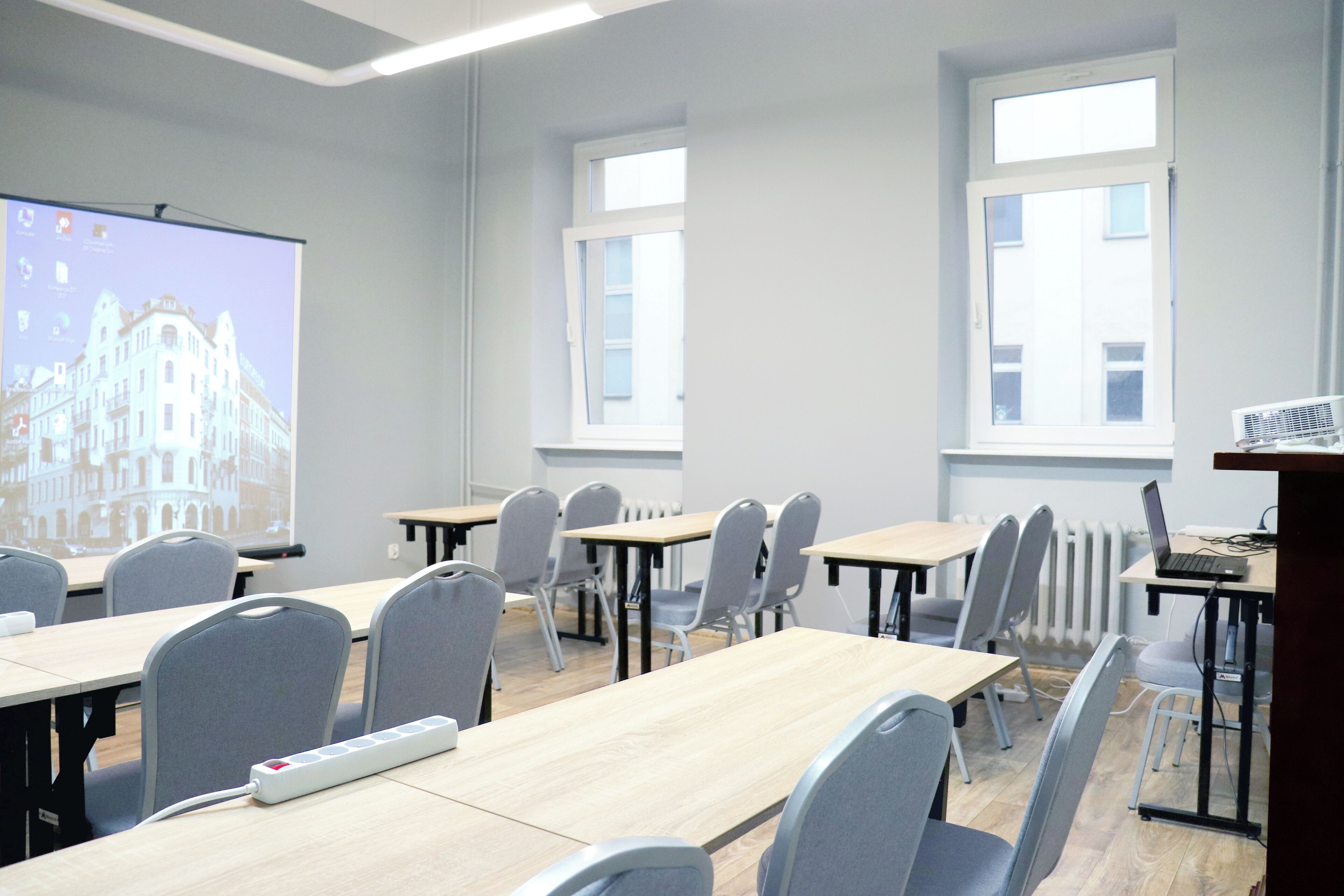 Uma sala de aula com mesas, cadeiras, um projetor e janelas.