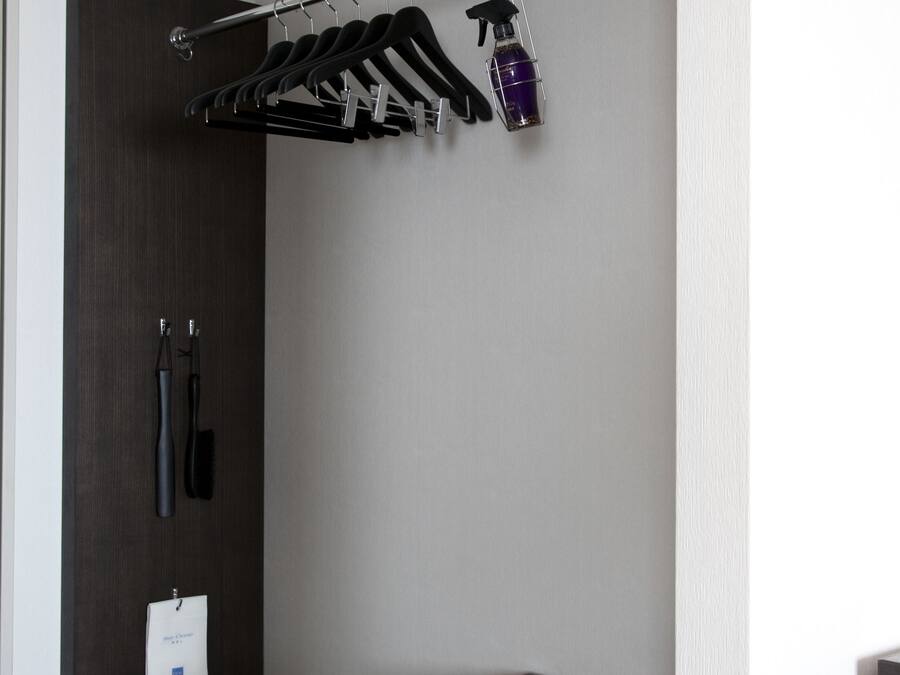 Quarto de hotel com uma estrutura metálica contendo vários cabides pretos, um borrifador e uma pequena lousa na parede. Há uma mesinha dobrável preta e um par de sapatos pretos no chão.
