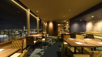 Quarto de hotel com vista para a cidade, área de bar e espaço de jantar com mesas e cadeiras.