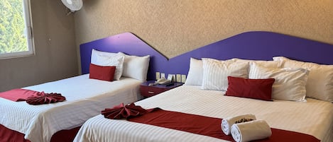 Quarto de hotel com duas camas, cada uma com roupa de cama vermelha e branca, um ventilador de teto e uma janela com vista para o verde.