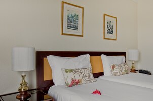 Quarto de hotel com duas camas, cabeceira de madeira, roupa de cama branca, almofada floral, objeto decorativo vermelho, mesa de cabeceira com abajur e duas fotos emolduradas na parede.