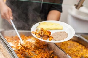 Uma pessoa servindo comida de um buffet, com um prato contendo um pedaço de omelete, cebolas fritas e um hambúrguer.