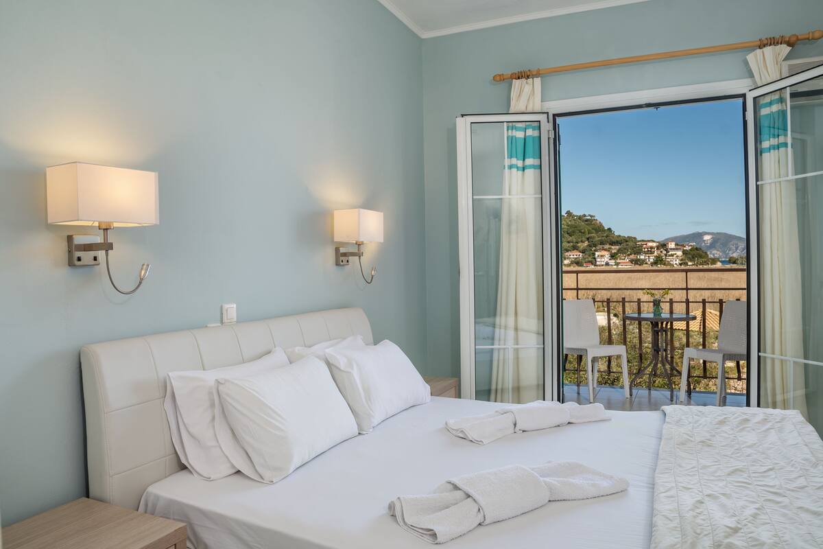Quarto de hotel com cama, duas luminárias fixadas na parede, mesa de cabeceira e varanda com mesa e cadeiras.