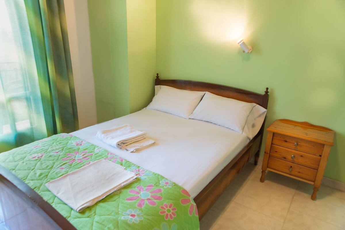 Cama bem arrumada com colcha floral verde e travesseiros brancos, mesa de cabeceira de madeira com gavetas, e luminária fixada na parede.
