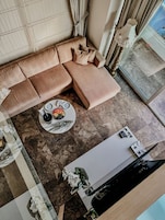 Uma sala de estar moderna com um sofá em formato de L, uma mesa de centro com frutas e bebidas, um aparador branco e uma porta de vidro que dá para uma varanda.