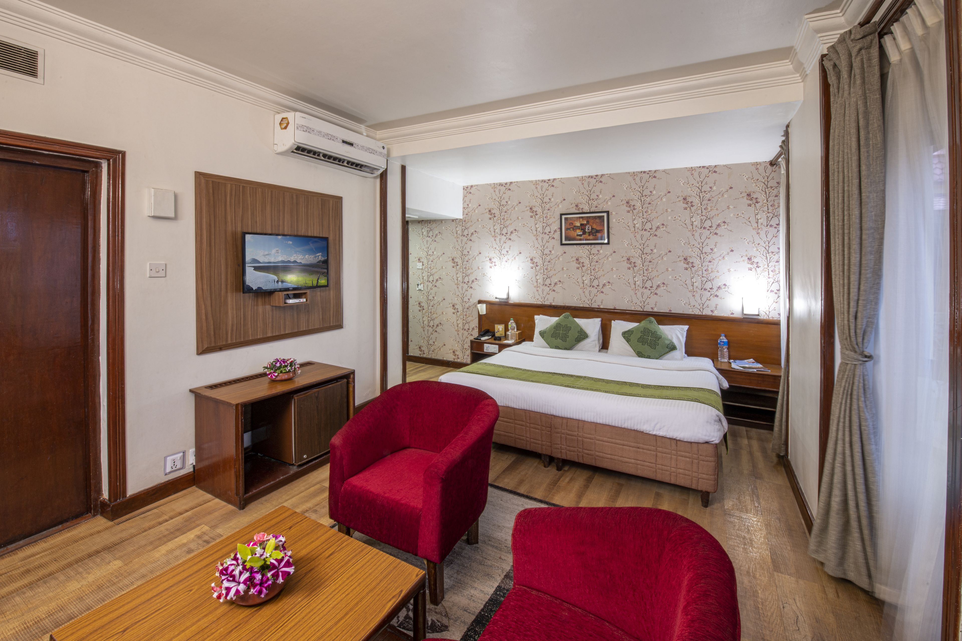 Quarto de hotel com uma cama, televisão, escrivaninha e duas cadeiras vermelhas.