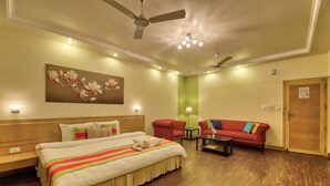 Quarto de hotel com uma cama grande, duas poltronas, uma mesa de centro, um ventilador de teto e uma porta.