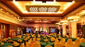 Uma sala de banquetes com mesas redondas cobertas por toalhas verdes e cadeiras brancas, um palco com uma banda, e teto decorativo.