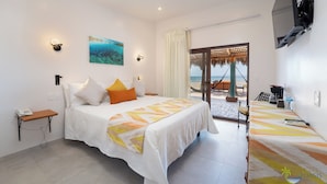Quarto de hotel com uma cama, uma escrivaninha com cadeira, uma televisão e vista para o mar através de uma porta de vidro.
