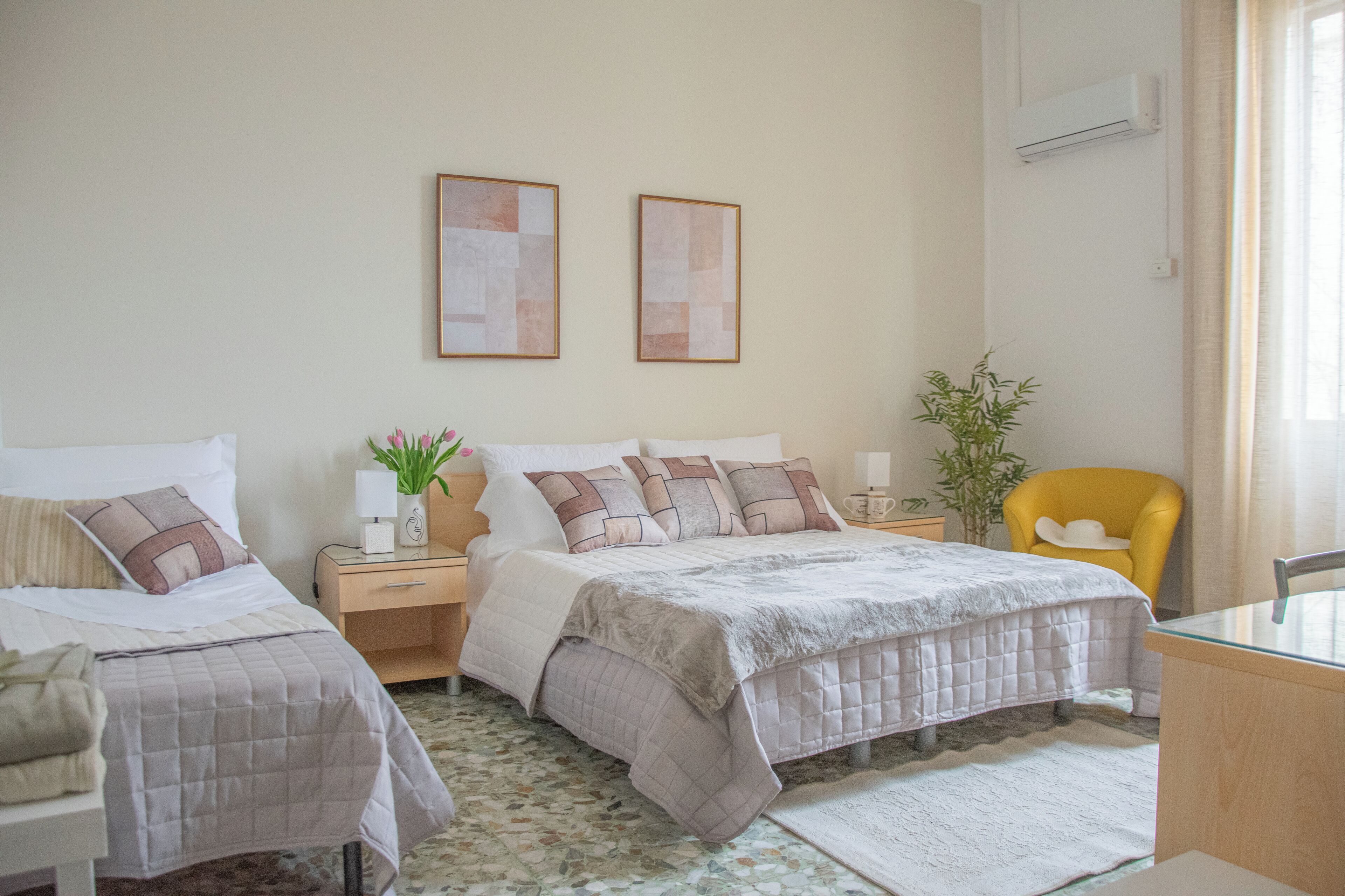 Quarto de hotel com duas camas, uma escrivaninha, uma cadeira, uma planta e obras de arte na parede.