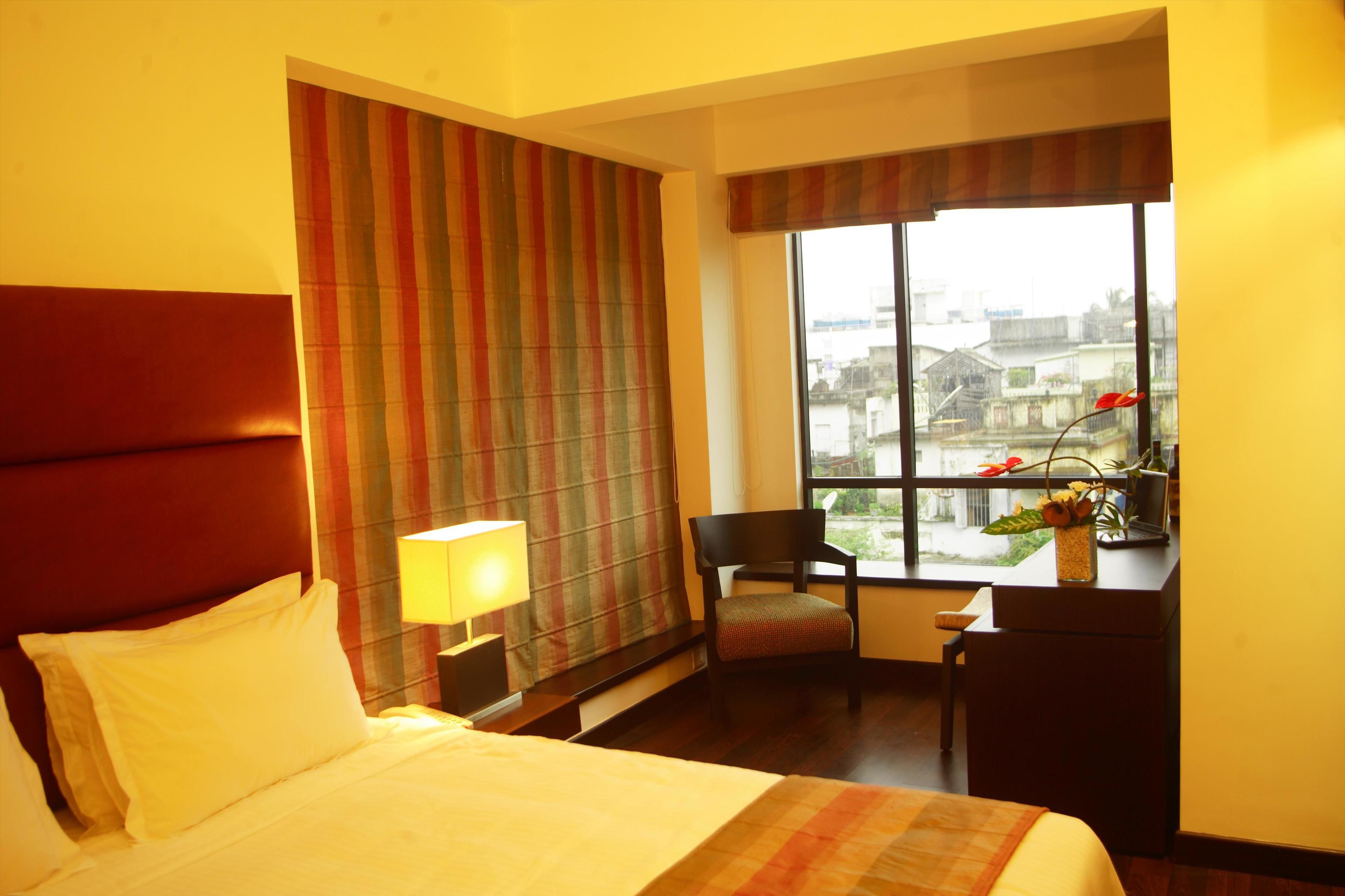 Quarto de hotel com cama, cadeira, escrivaninha, abajur e janela com vista para edifícios.