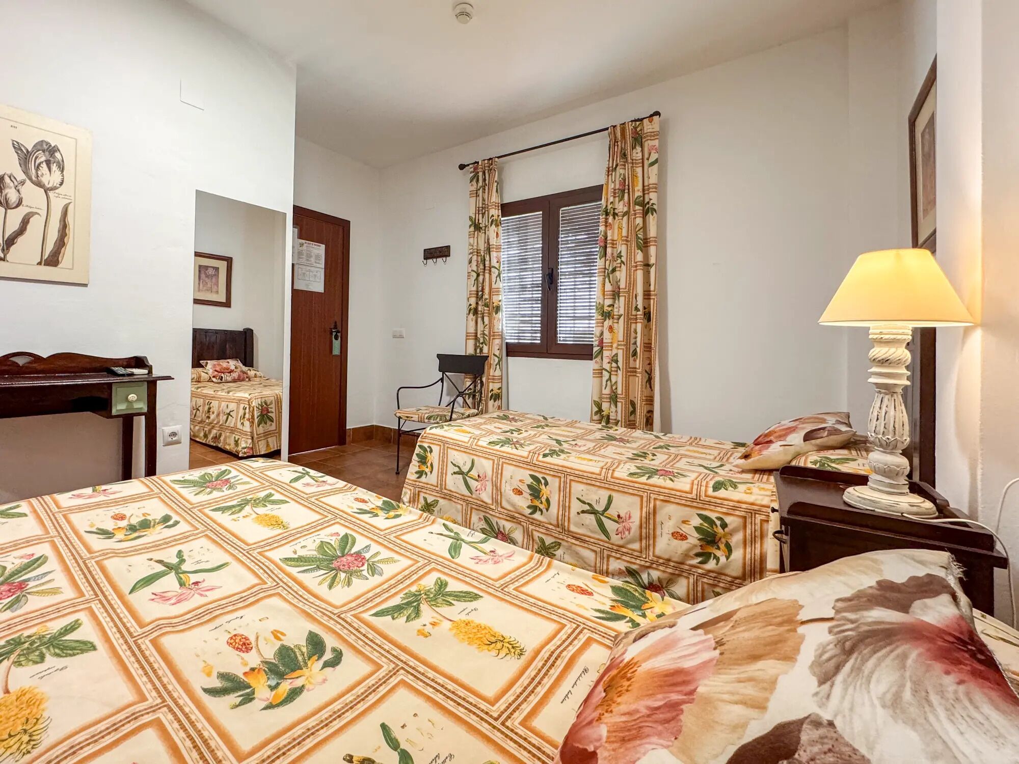 Quarto de hotel com duas camas, uma escrivaninha, uma cadeira, um abajur e uma janela com cortinas.