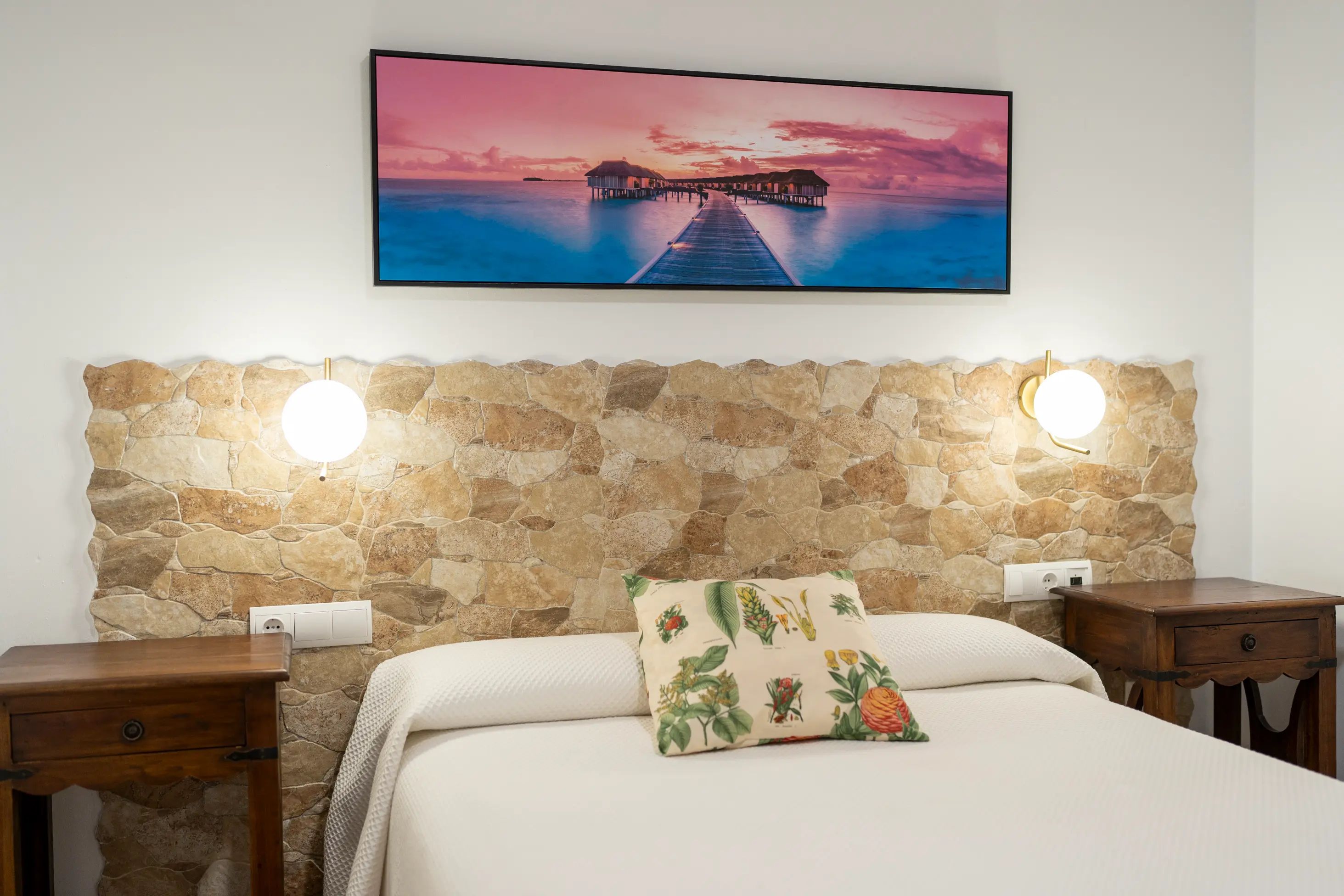 Cama com travesseiro floral, parede de pedra, duas mesas de cabeceira e pintura com vista para o pôr do sol.