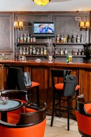 Uma área de bar com painéis de madeira, uma televisão montada acima e diversos tipos de bebidas. Há cadeiras laranja e uma mesa com um copo e uma flor.
