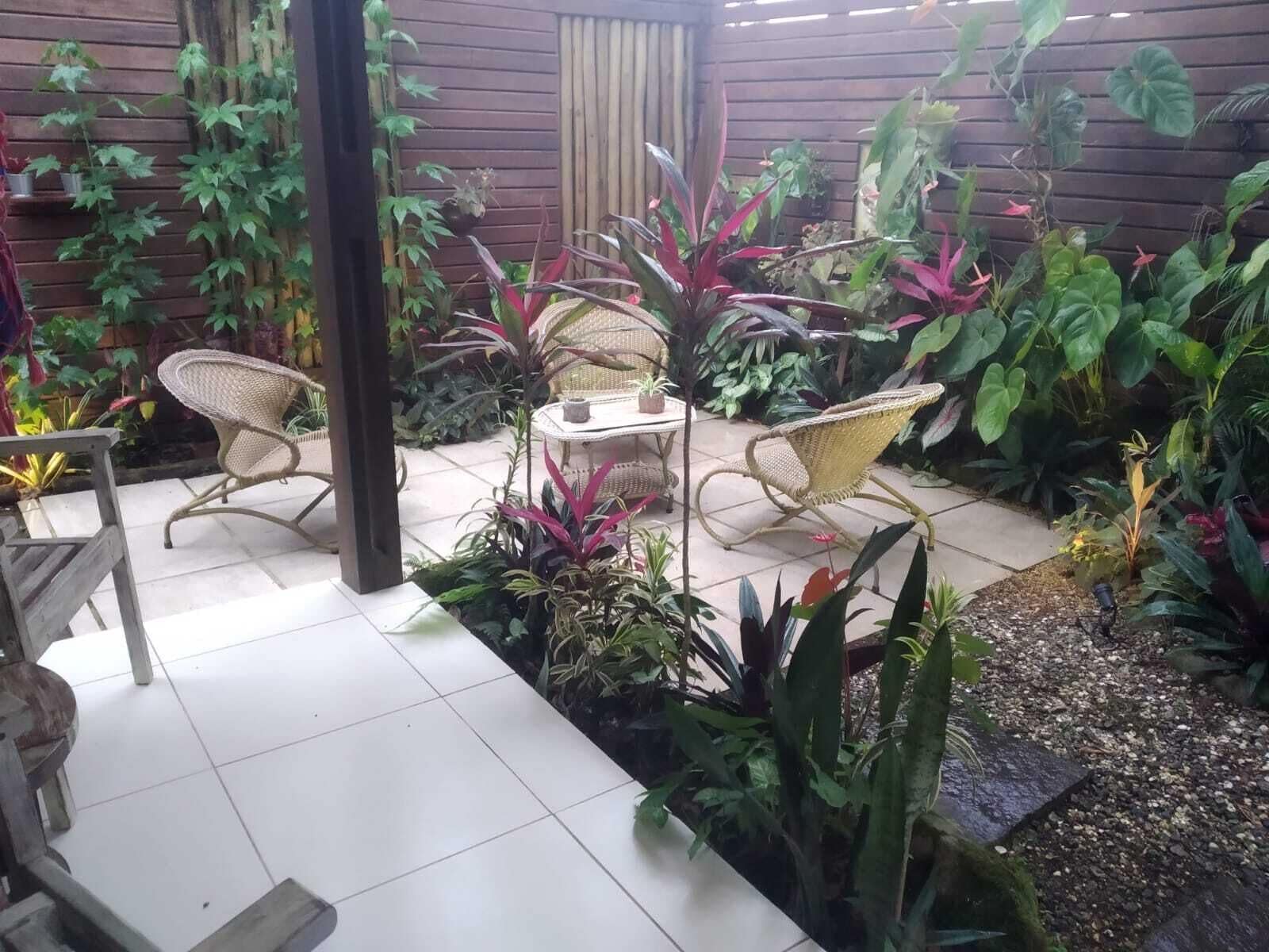 Uma área de pátio com móveis de vime, piso de azulejos e diversos vasos de plantas.