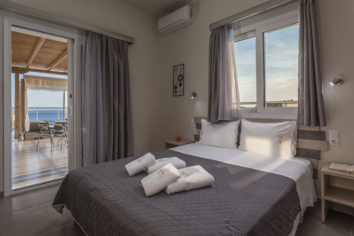Quarto de hotel com cama, mesa de cabeceira, janela com cortinas, vista para o mar e varanda com cadeiras.