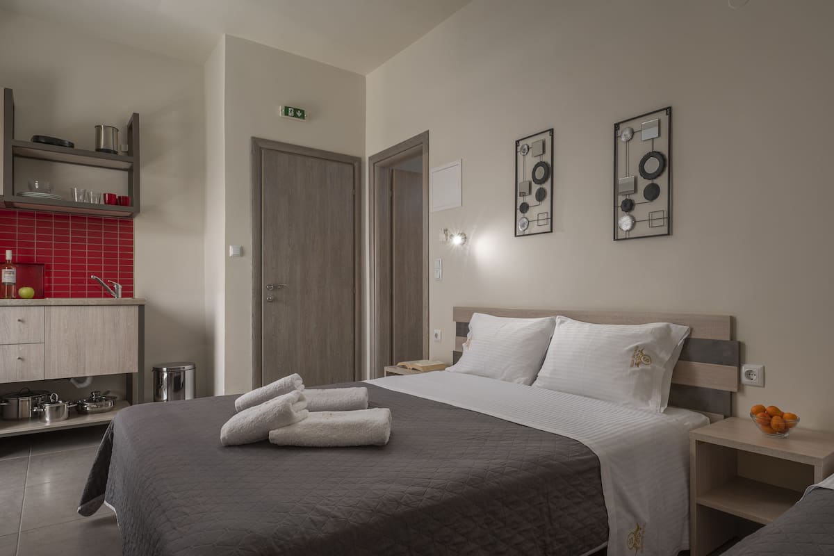 Quarto de hotel com cama, mesa de cabeceira e uma pequena mesa com laranjas. Há uma área de cozinha com painel vermelho e armários.