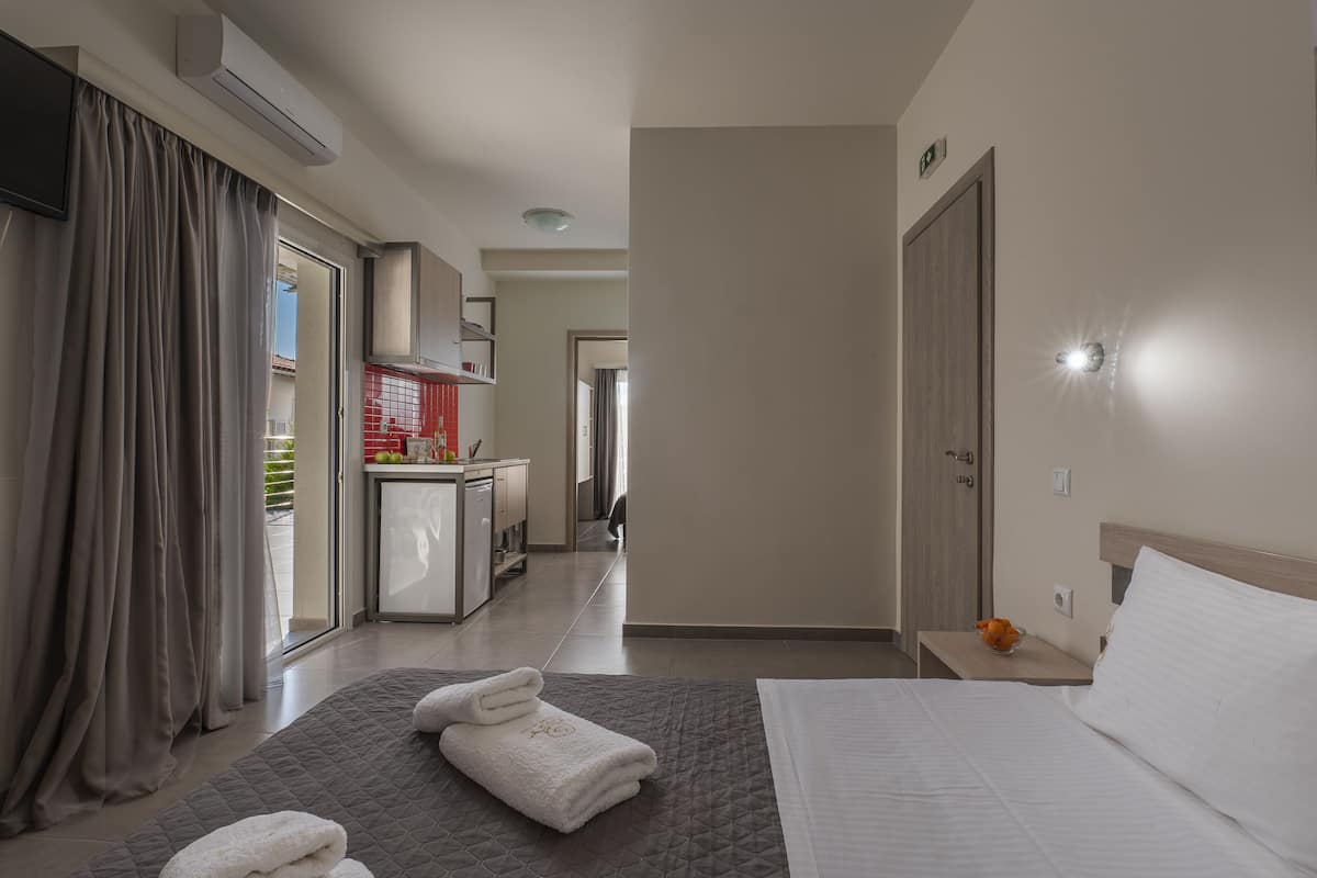 Quarto de hotel com cama, mesa de cabeceira com uma cesta de frutas, escrivaninha com computador e uma cozinha compacta ao fundo.