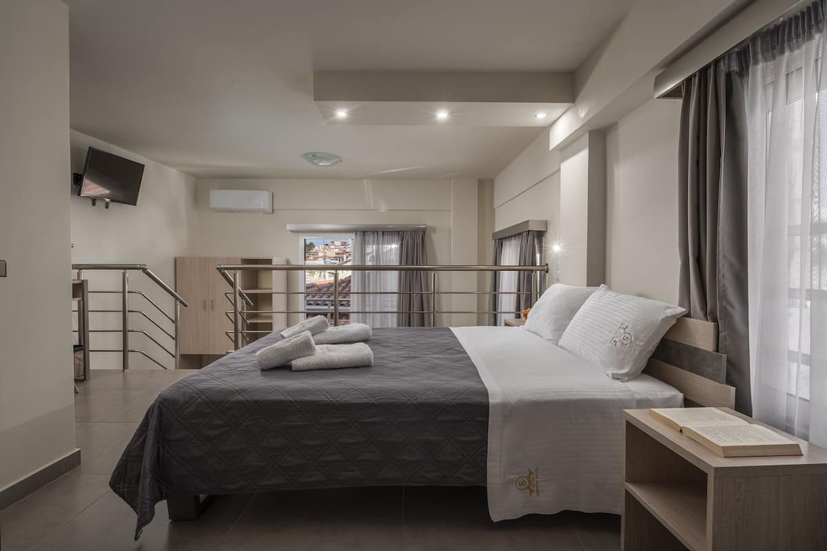 Quarto de hotel moderno com uma cama grande, uma mesinha de cabeceira com um livro, uma TV na parede e vista para o exterior pela janela.
