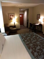 Quarto de hotel com cama, escrivaninha, duas cadeiras, abajur e porta que dá para outro espaço.