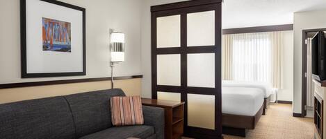 Quarto de hotel com sofá, mesa pequena, cama, televisão e um quadro na parede.