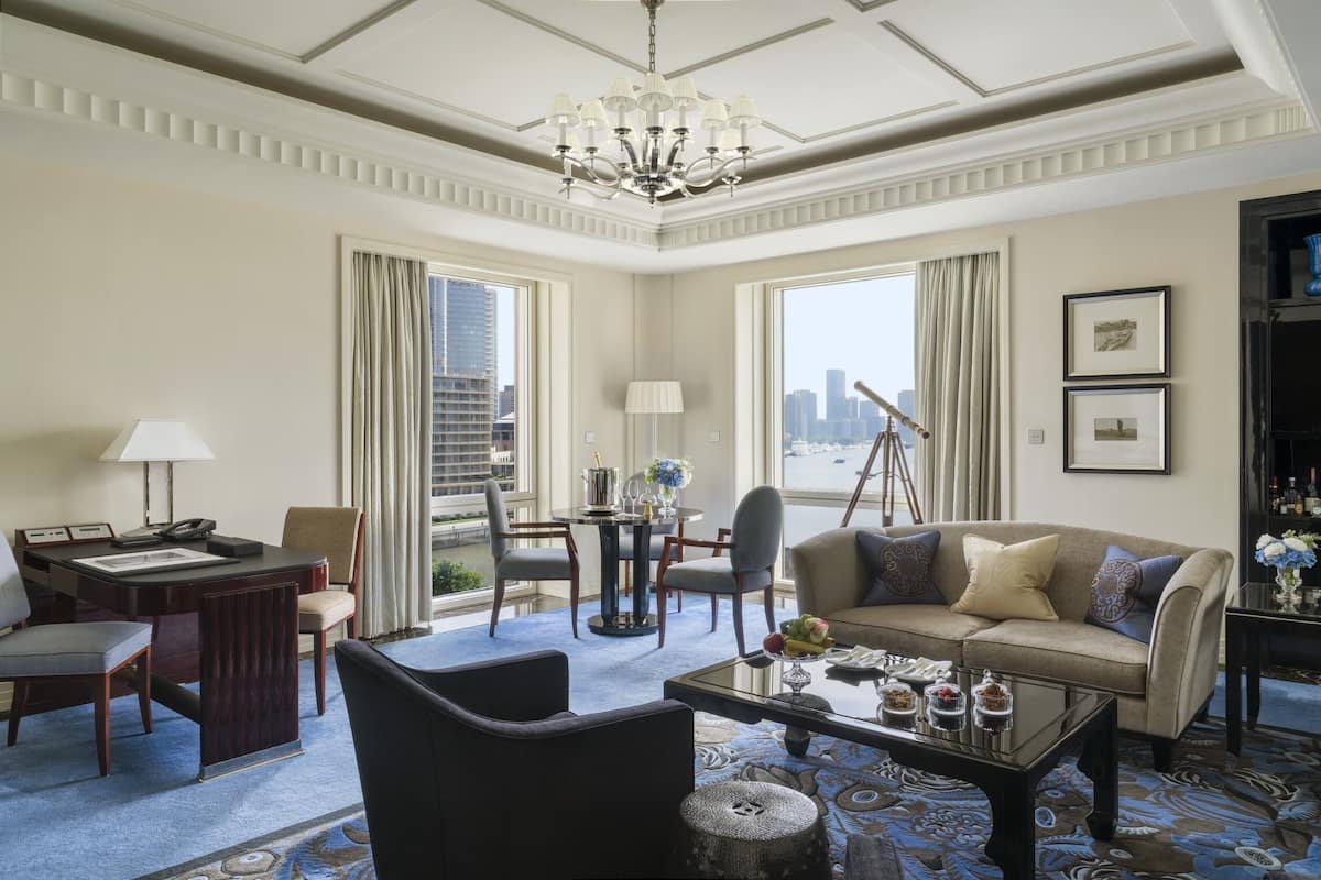 Quarto de hotel com uma grande janela, um candelabro, um sofá, uma mesa de centro e uma escrivaninha com uma luminária.