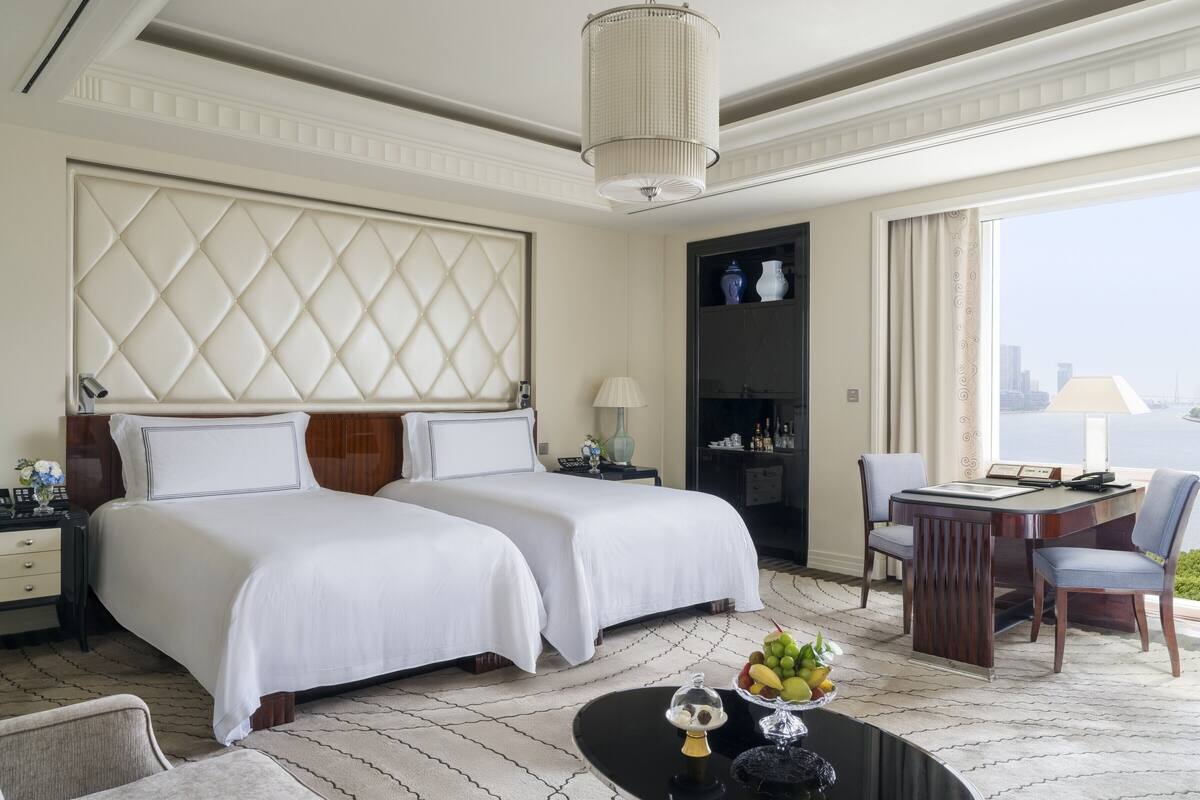 Quarto de hotel com duas camas, uma escrivaninha, uma cadeira, vista da cidade e uma janela grande.