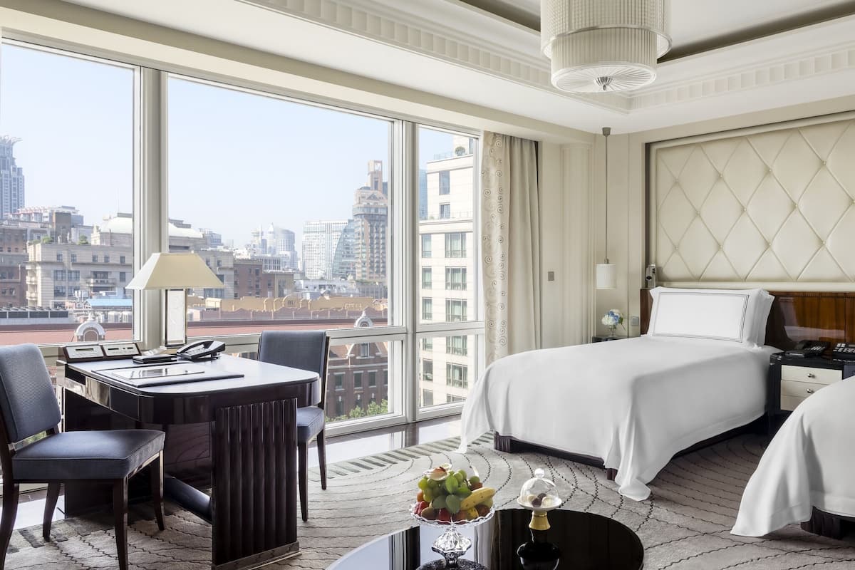 Quarto de hotel com uma janela grande, uma escrivaninha com cadeira, duas camas e uma pequena mesa com um vaso de flores.