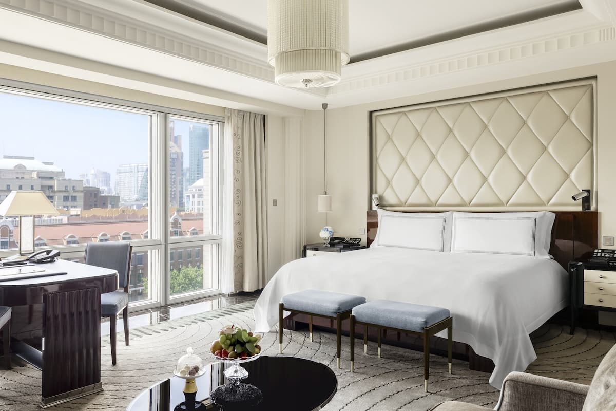 Quarto de hotel com uma cama grande, uma escrivaninha e uma cadeira. Vê-se a cidade através de grandes janelas.