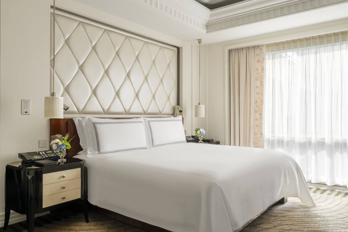 Quarto de hotel com uma cama grande, mesa de cabeceira com abajur, telefone e um vaso com flores.