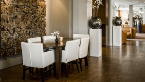 Uma sala de jantar com parede de pedra, cadeiras brancas, mesa de madeira e grandes vasos com flores.