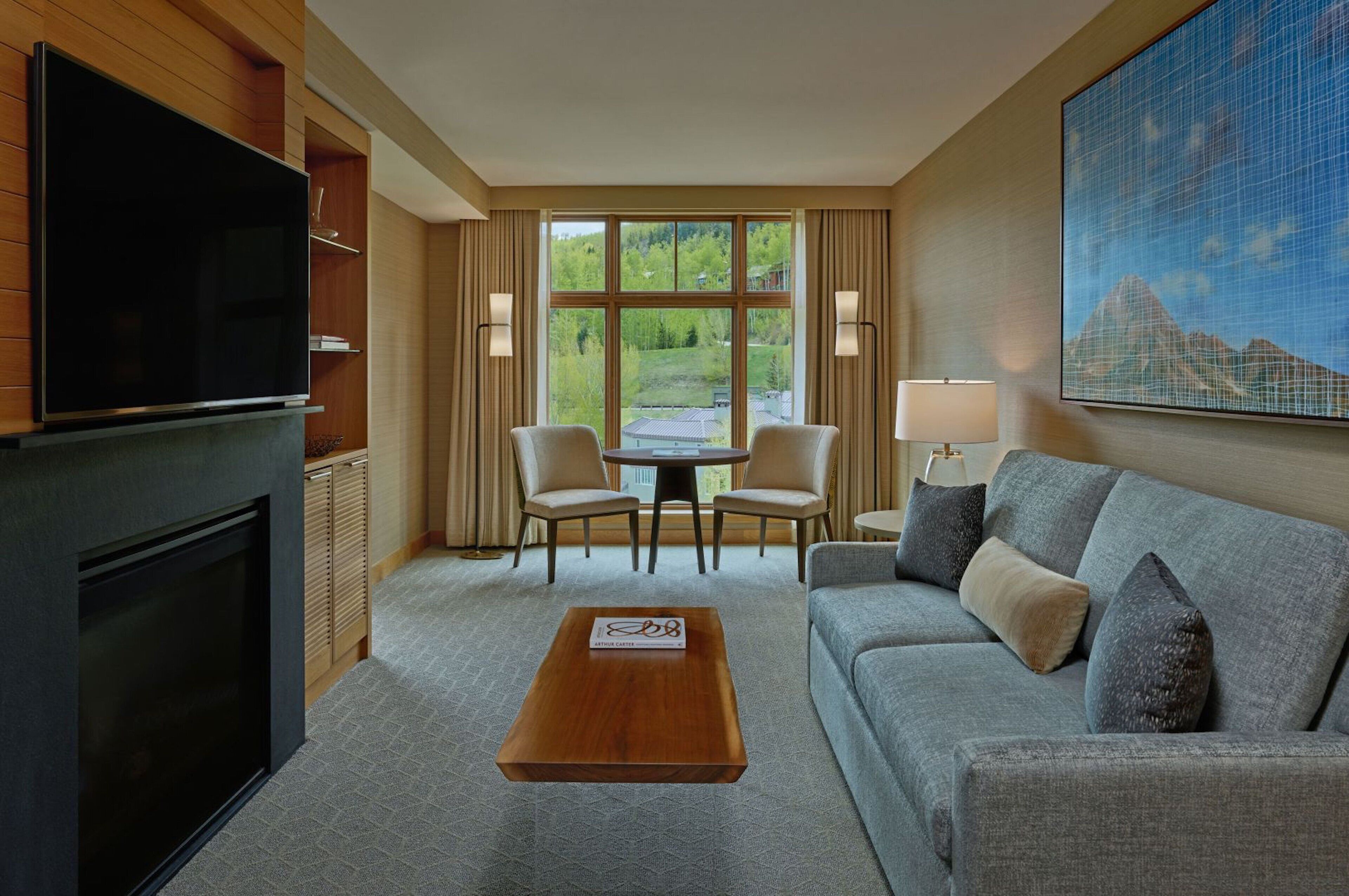 Quarto de hotel moderno com televisão de tela plana, lareira, sofá, mesa de centro e uma grande janela com vista para o verde.