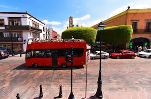 Um ônibus de dois andares vermelho estacionado em uma praça com calçada de tijolos, cercado por edifícios e uma torre do relógio ao fundo.