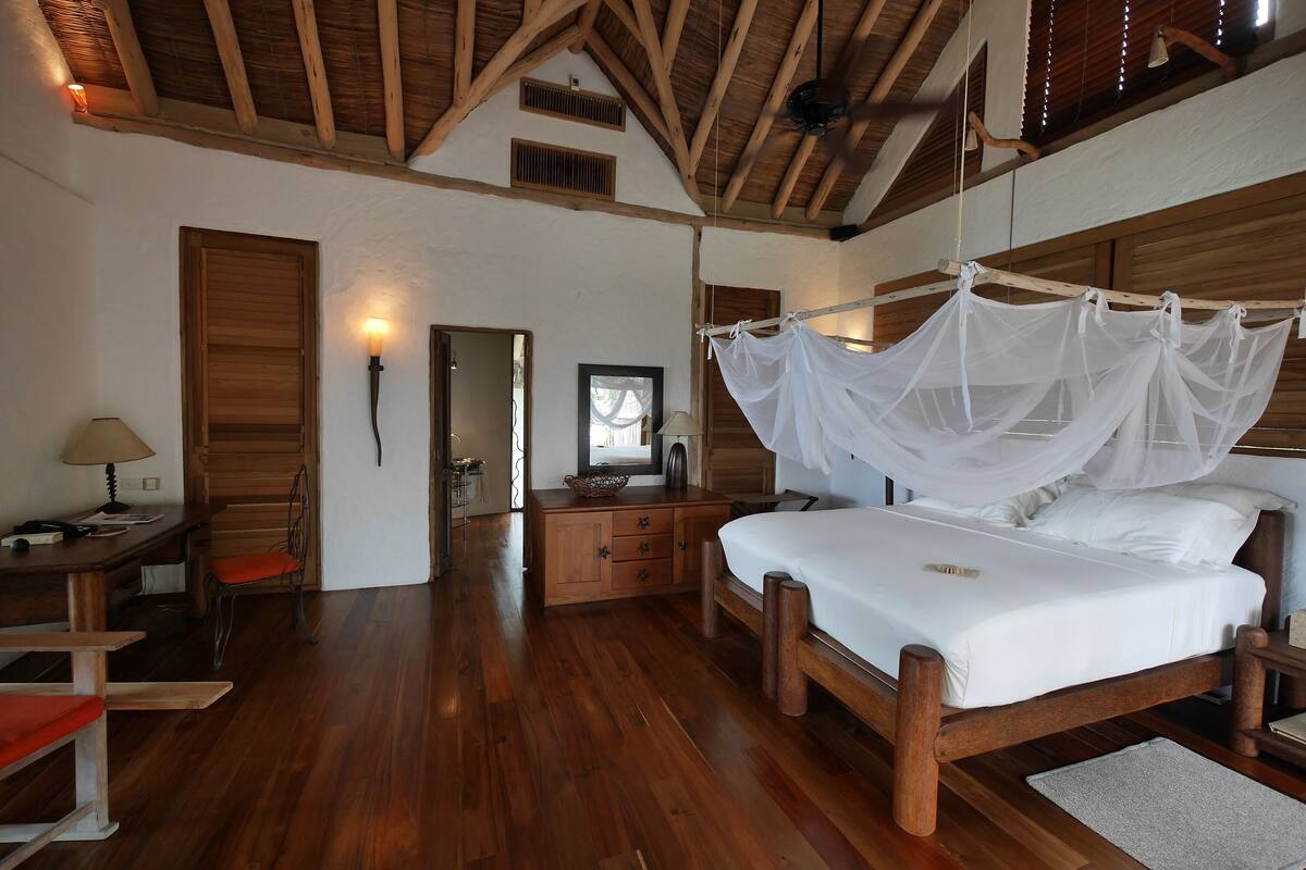 Quarto com cama de madeira, mesa de cabeceira, escrivaninha com cadeira e espelho.
