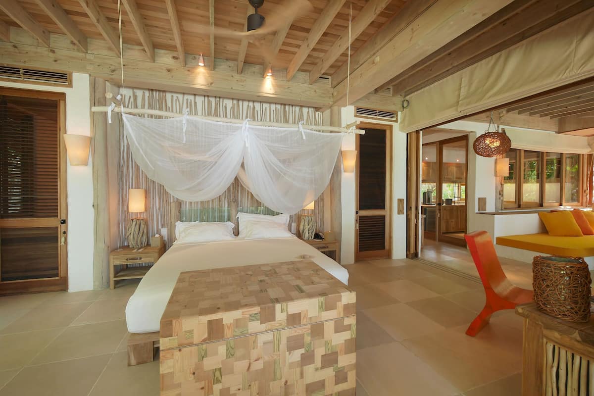Quarto com cama grande, cabeceira de madeira e dossel. Há uma mesa de cabeceira de madeira com abajur e cesta de vime.