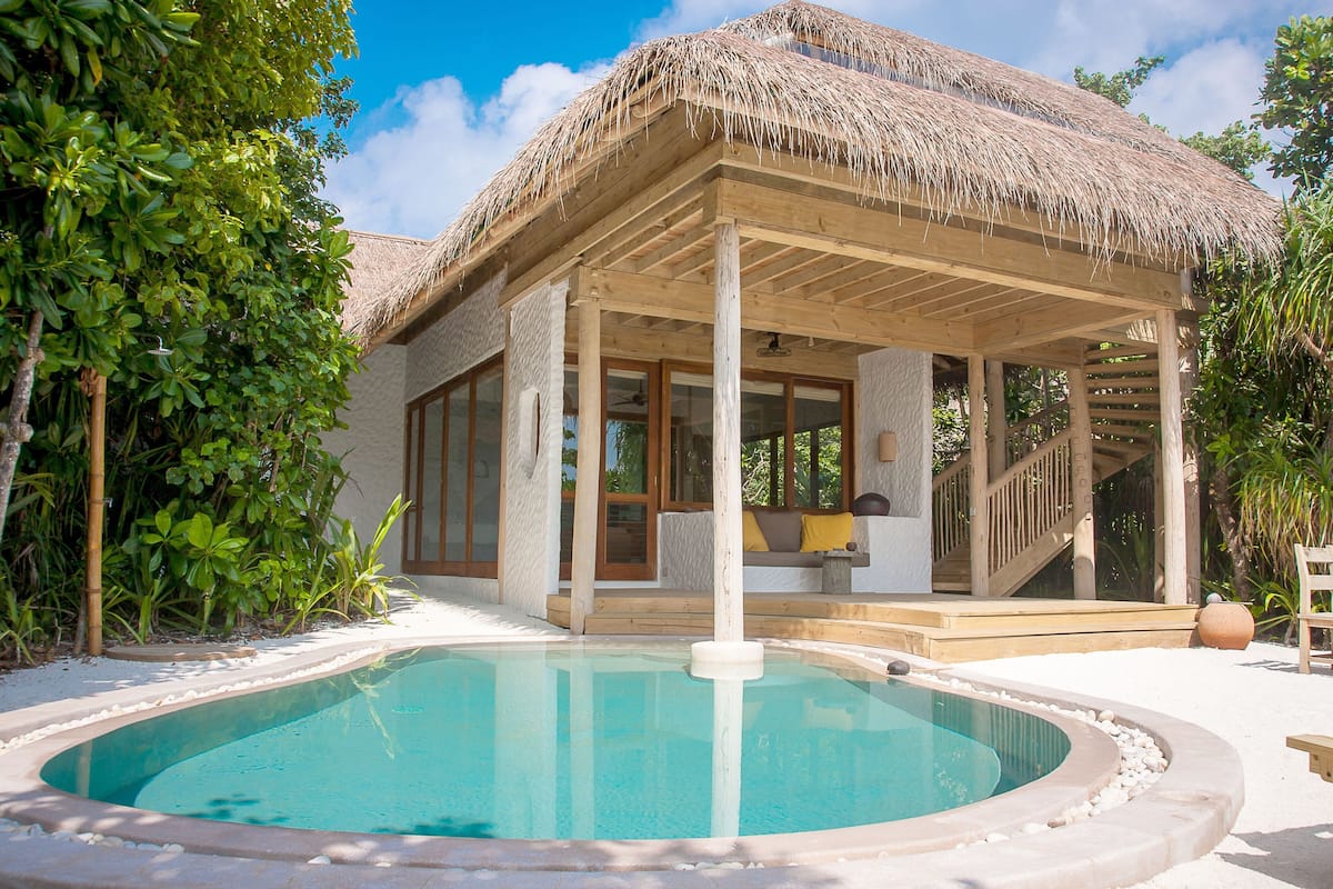 Uma área ao lado da piscina com uma estrutura de telhado de palha, piscina e vegetação ao redor.