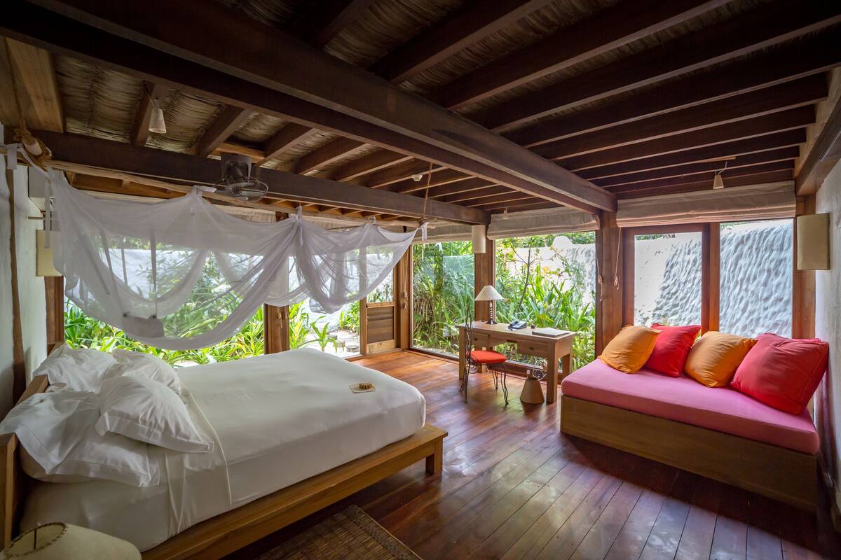 Quarto com cama, sofá, escrivaninha e janela com vista para o verde.