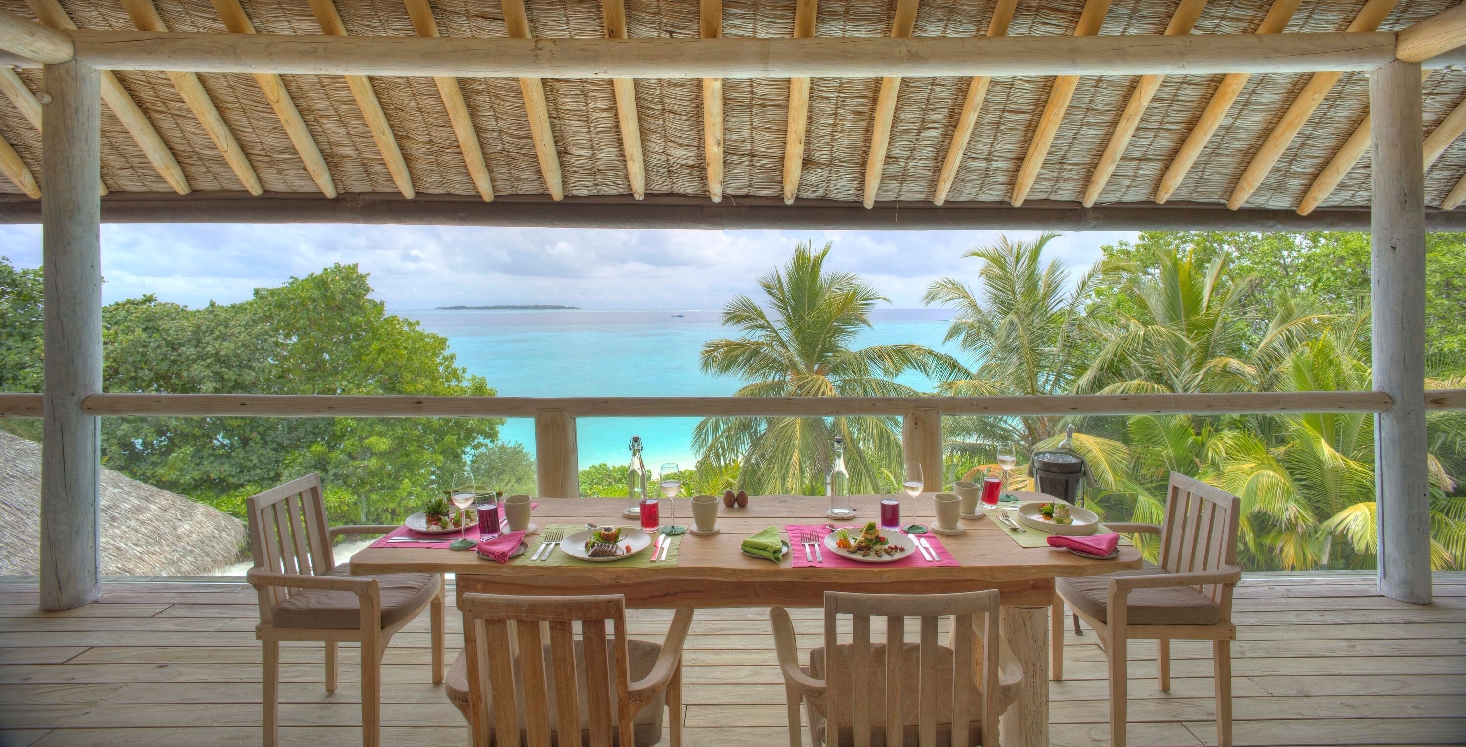 Um deck de madeira com uma mesa posta para refeição, com vista para uma praia tropical e oceano.