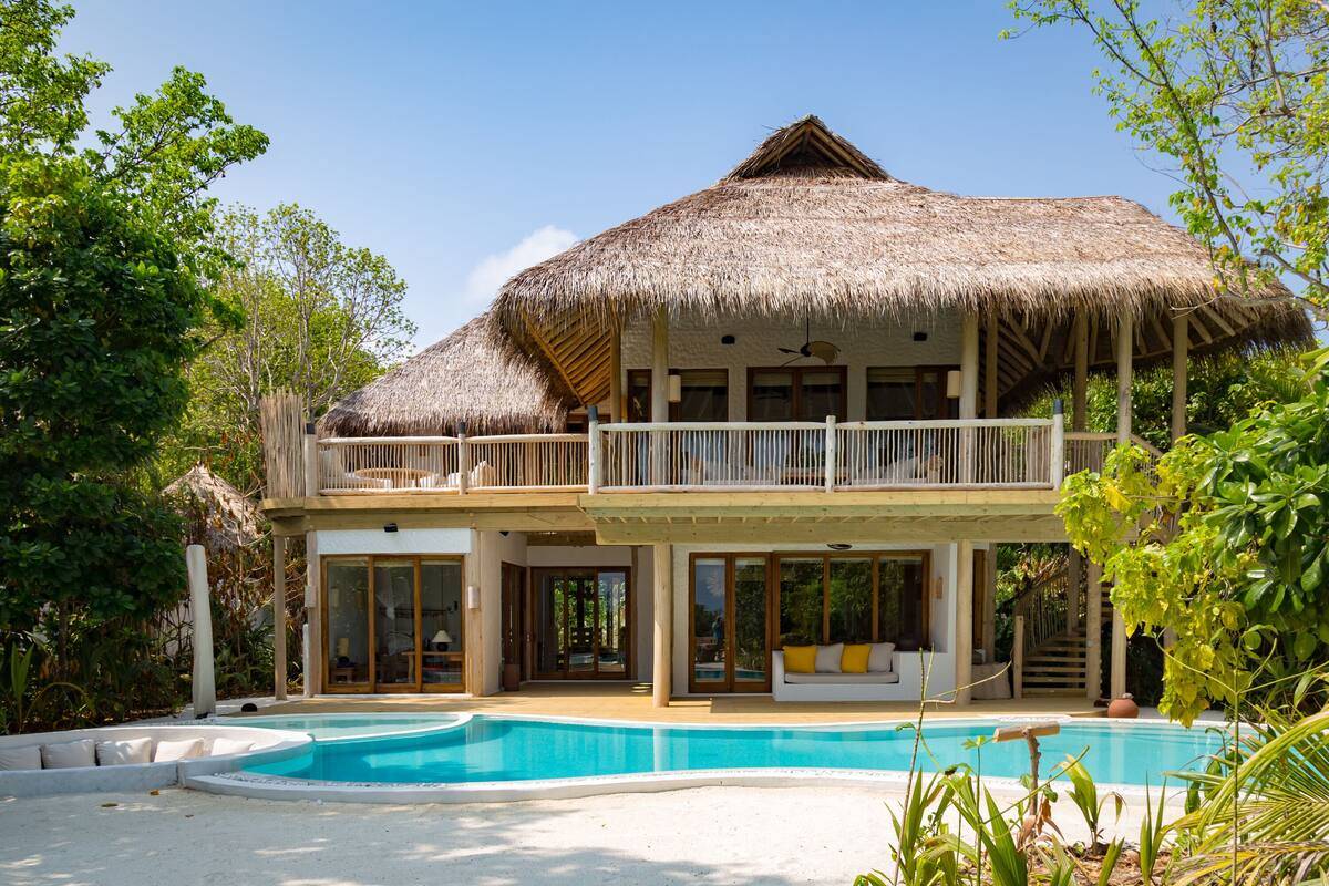 Uma casa de dois andares com telhado de palha, piscina e varanda.