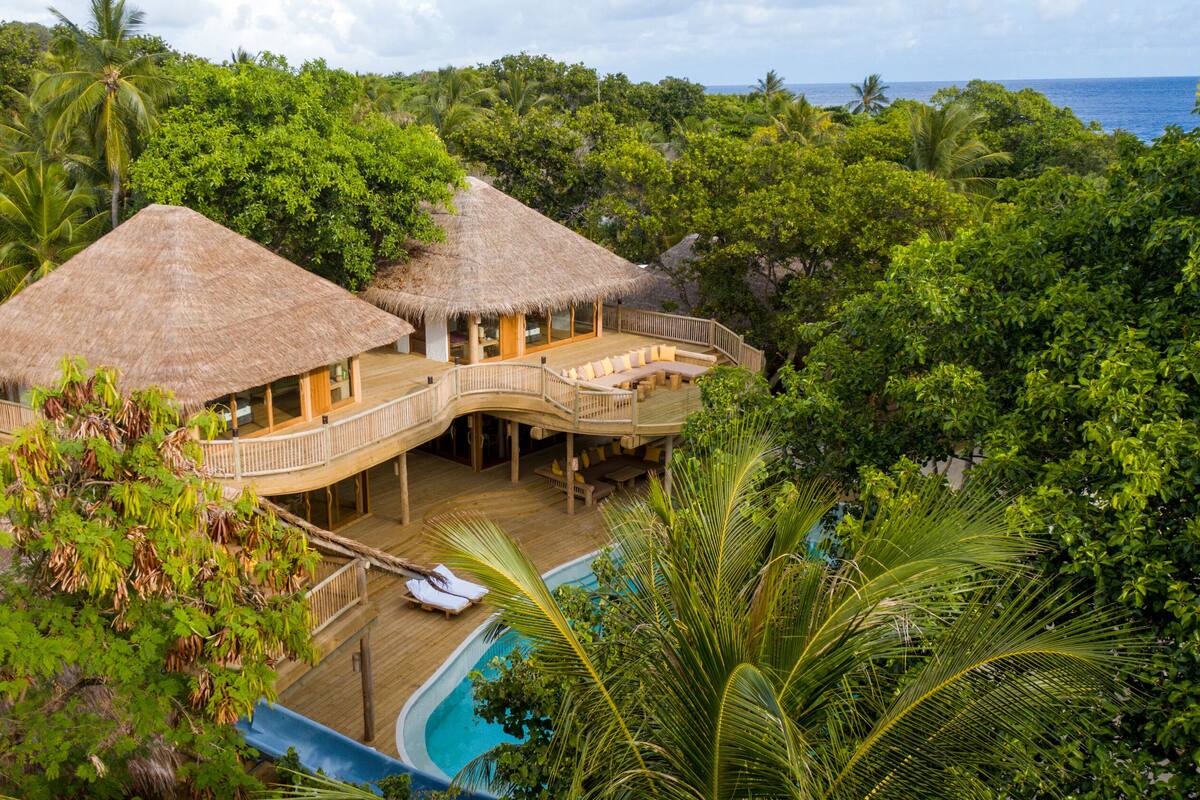 Resort com duas estruturas de telhado de palha interligadas por uma ponte de madeira, piscina e vegetação tropical exuberante.