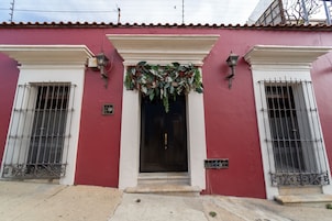 Um edifício vermelho com porta branca, duas janelas com grades de metal e uma coroa decorativa sobre a porta.