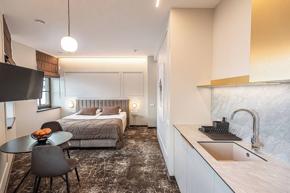 Quarto de hotel compacto com cama, mesa de cabeceira e pequena cozinha.