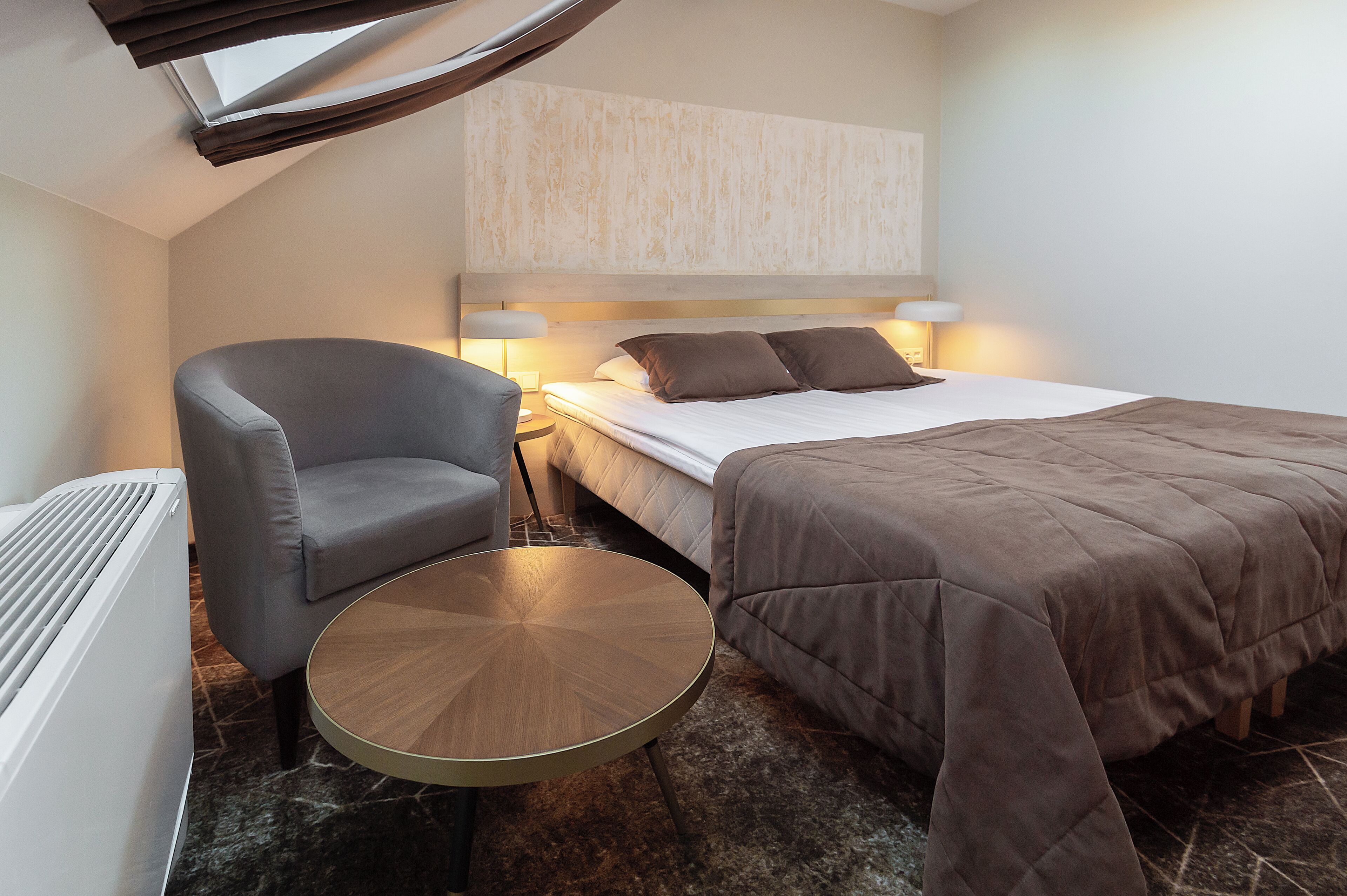 Quarto de hotel com cama, poltrona, mesa redonda e radiador.