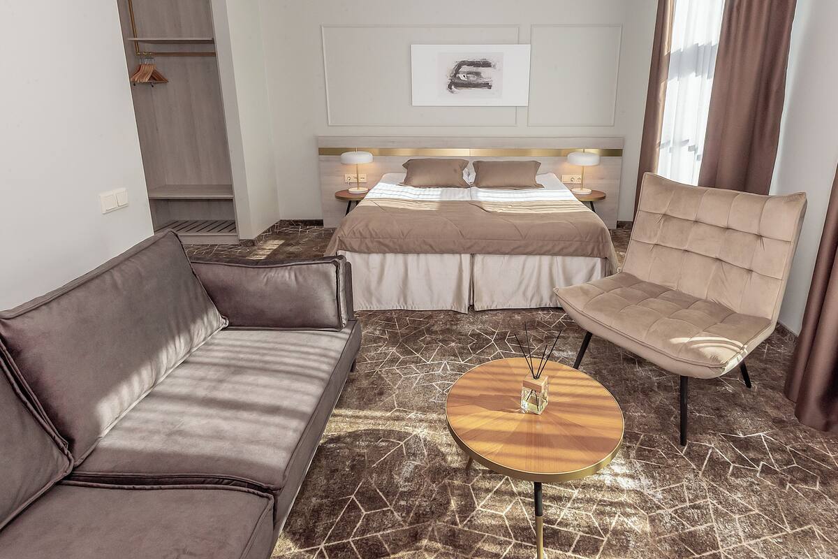 Quarto de hotel com cama, sofá, cadeira, mesa redonda e piso estampado.