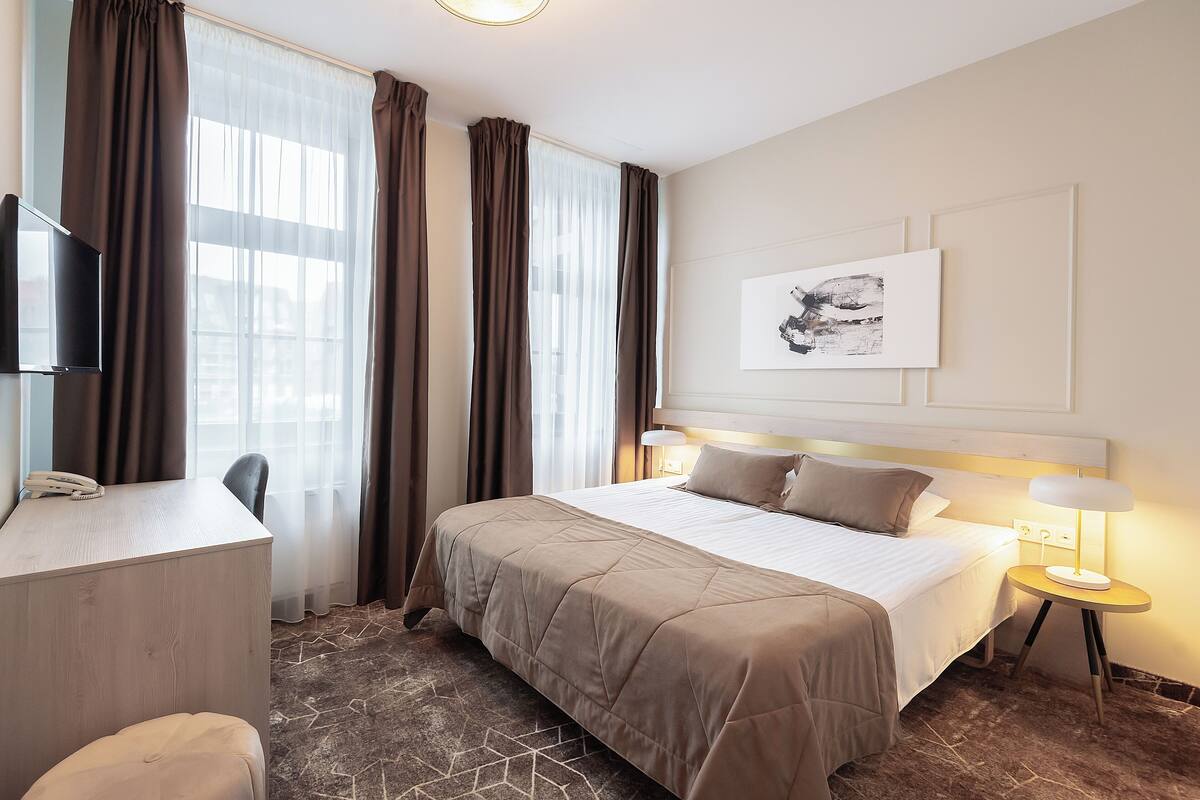 Quarto de hotel com cama, escrivaninha, cadeira, abajur e um quadro na parede.