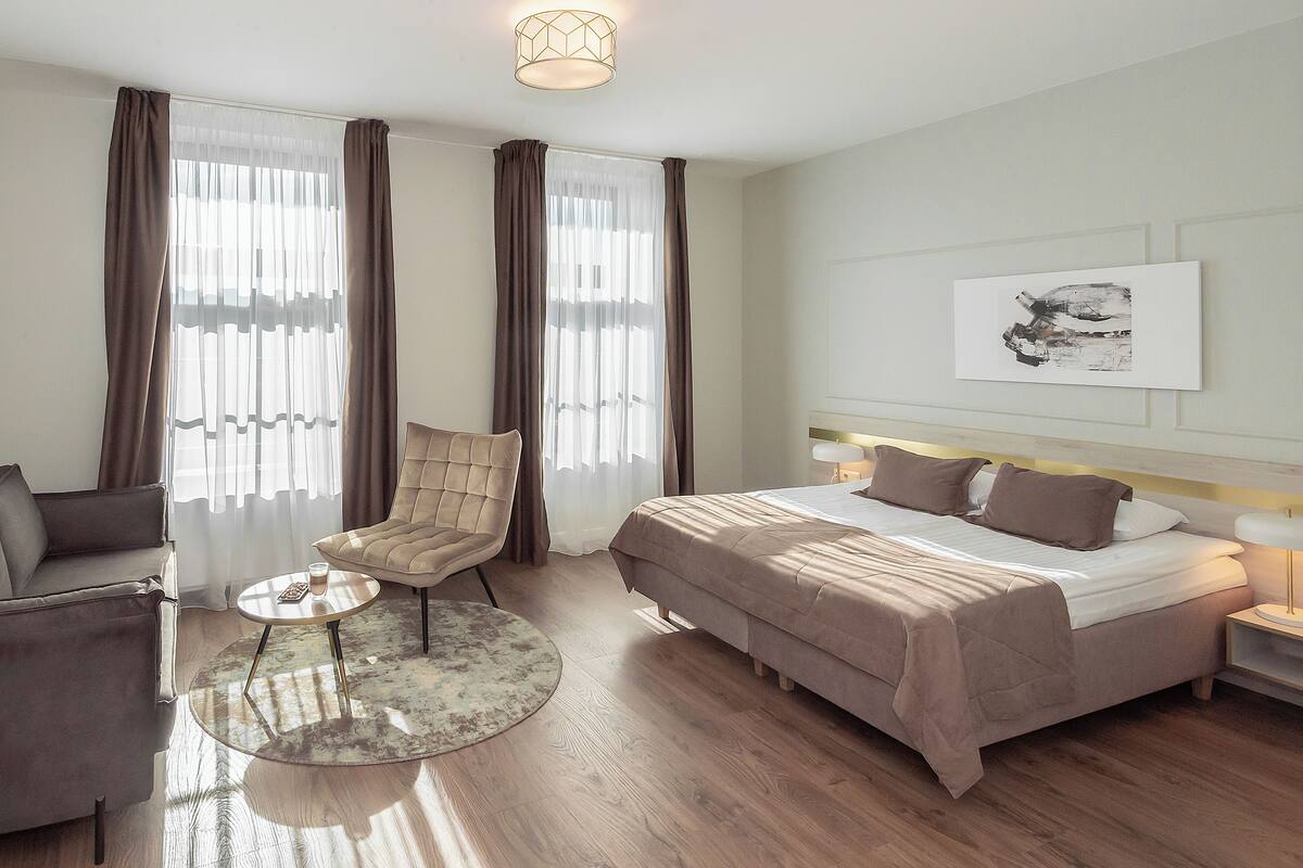 Quarto de hotel moderno com cama, sofá, poltrona, mesa pequena e um quadro na parede.