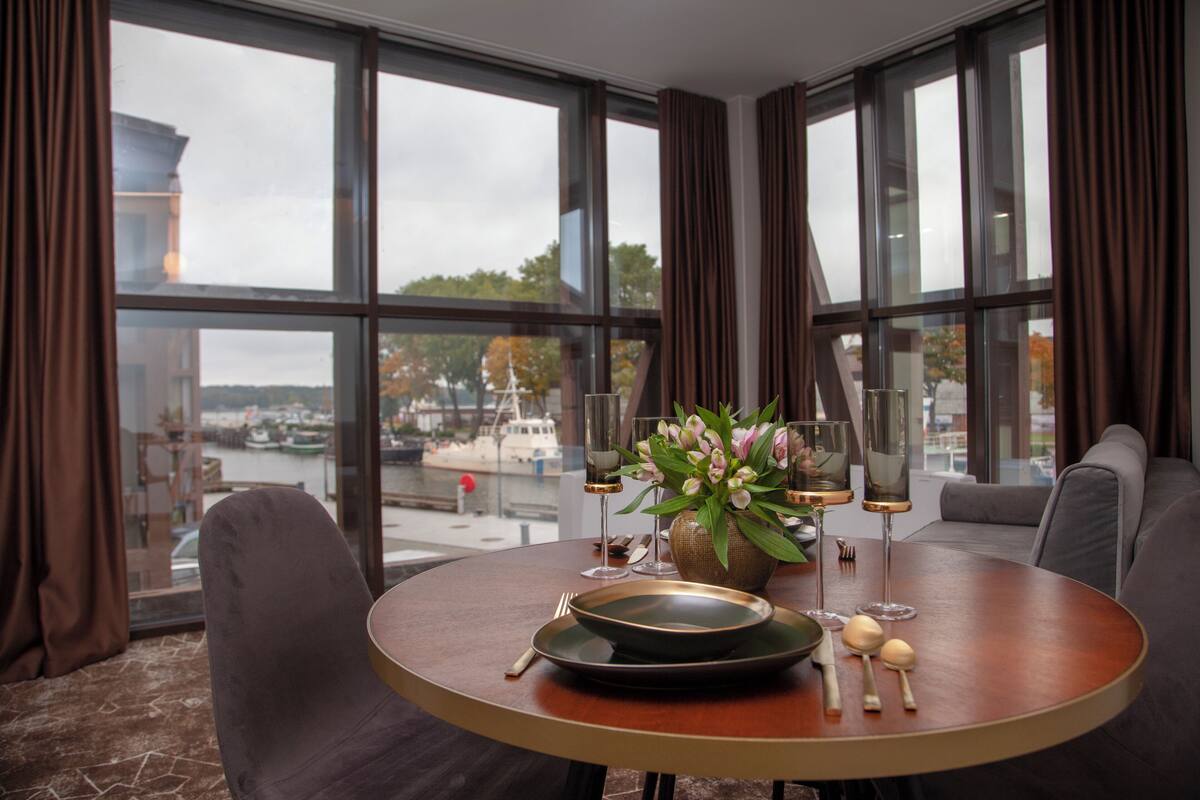 Uma mesa de jantar com um vaso de flores, duas taças de vinho e talheres, com uma vista panorâmica por grandes janelas para a água e edifícios.