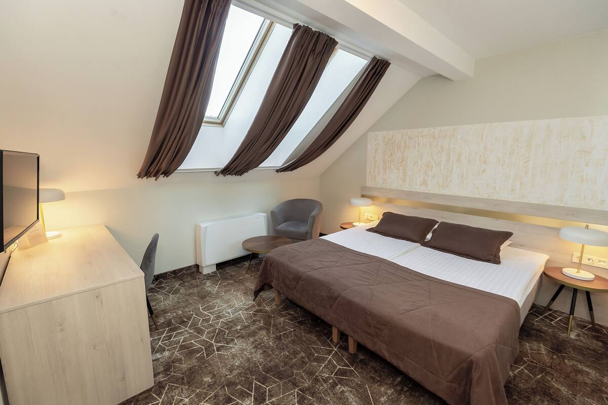 Quarto de hotel com cama, escrivaninha, cadeira, mesinha e janela com cortinas.