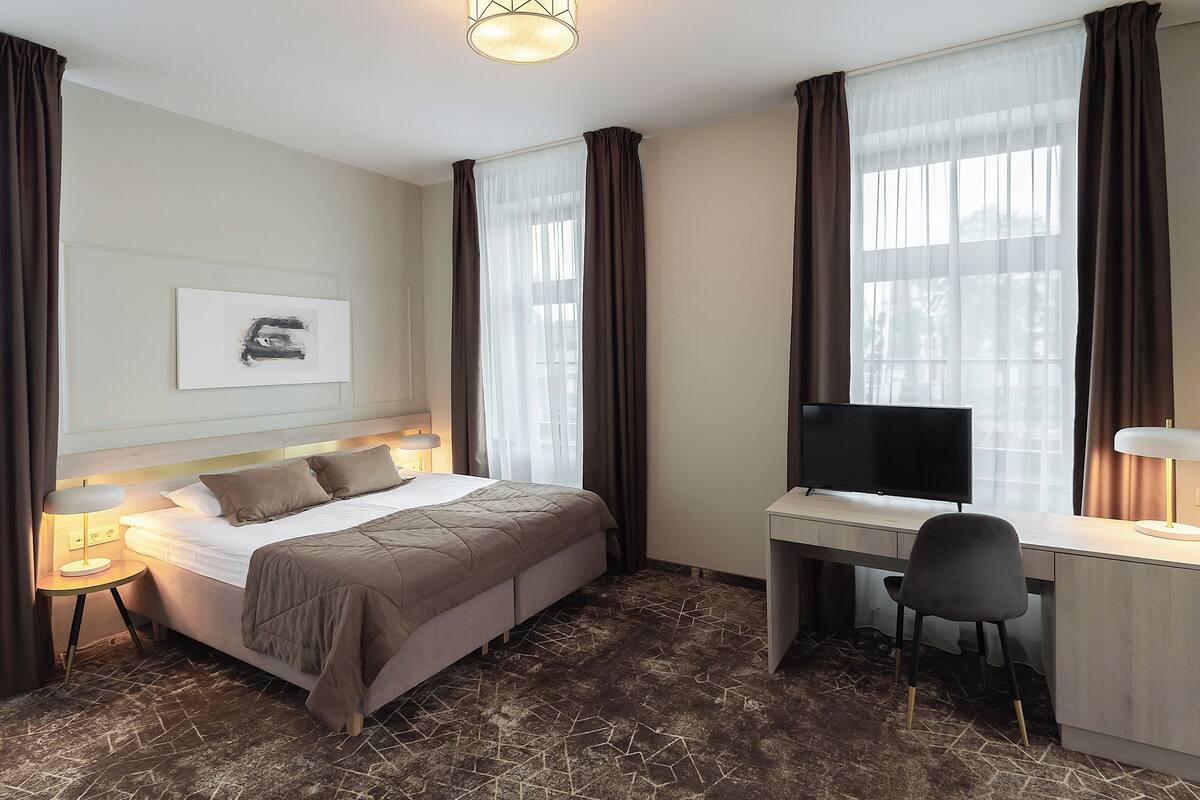 Quarto de hotel com cama, escrivaninha, cadeira e televisão.