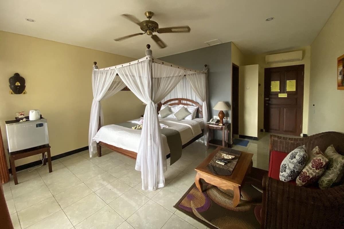 Quarto com cama de dossel, ventilador de teto, mesa de cabeceira com abajur, mesa de centro e sofá.